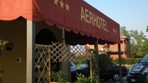 Exterior do Aerhotel com toldo vermelho, cerca treliçada e carros estacionados.