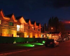 Uma fileira de edifícios de madeira com telhados inclinados, provavelmente um hotel ou resort, à noite, com iluminação pública e um carro estacionado na frente.