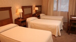 Quarto de hotel com três camas de solteiro, cada uma com cabeceira de madeira e mesa de cabeceira com luminária.