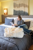 Uma pessoa sentada em uma cama com travesseiro, cobertor e uma bolsa branca.