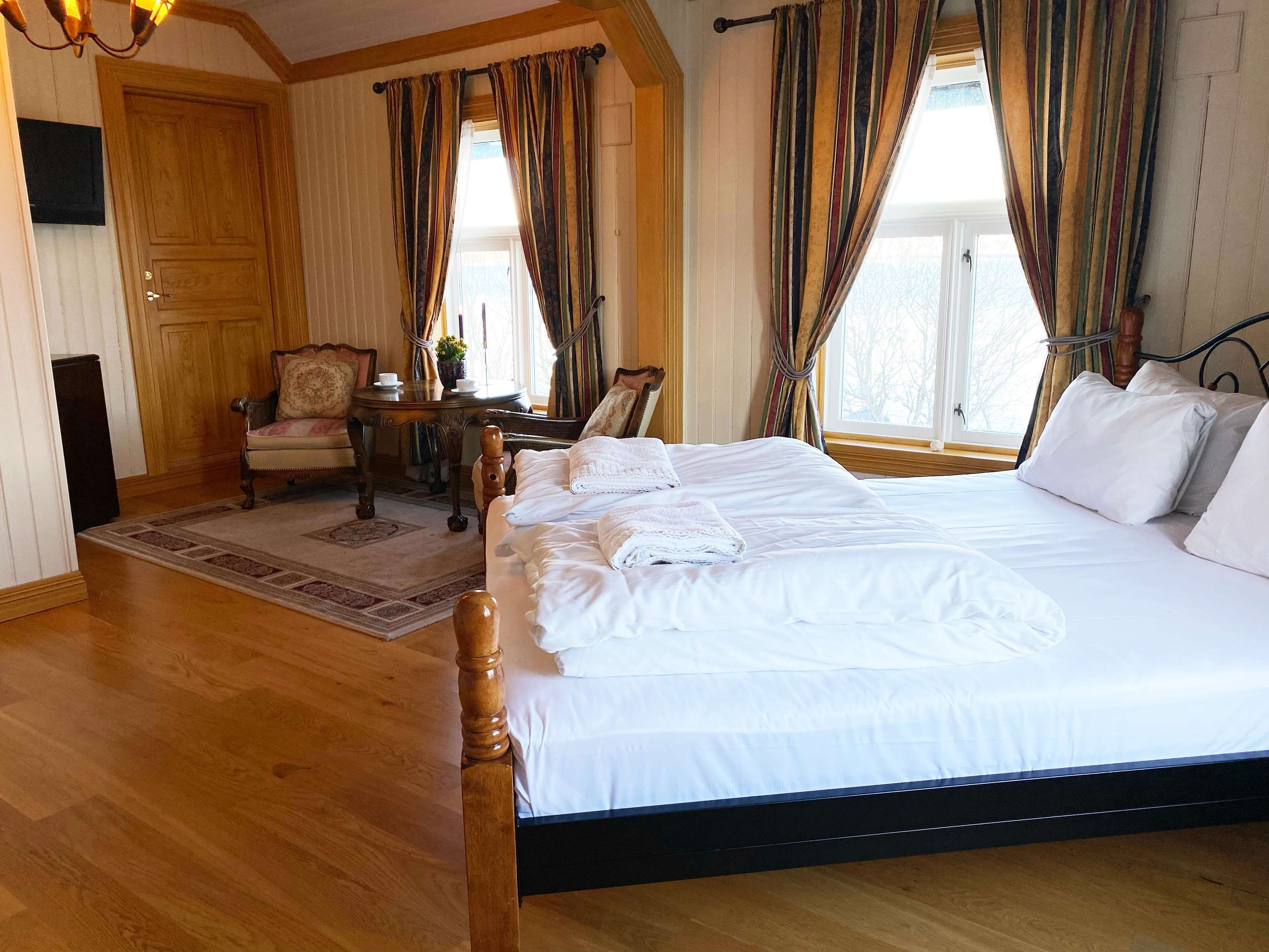Quarto com cama grande, piso de madeira, porta de madeira, uma mesinha com cadeira e cortinas.