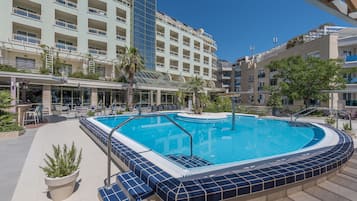 Piscina com deck ladrilhado, cercada por espreguiçadeiras e plantas em vasos.