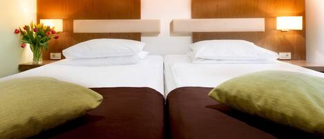 Quarto de hotel com duas camas, cada uma com roupa de cama branca e cabeceira marrom. Há duas luminárias de mesa e um vaso com flores na mesinha de cabeceira.
