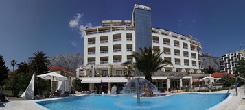 Um hotel com piscina, palmeiras e uma montanha ao fundo.