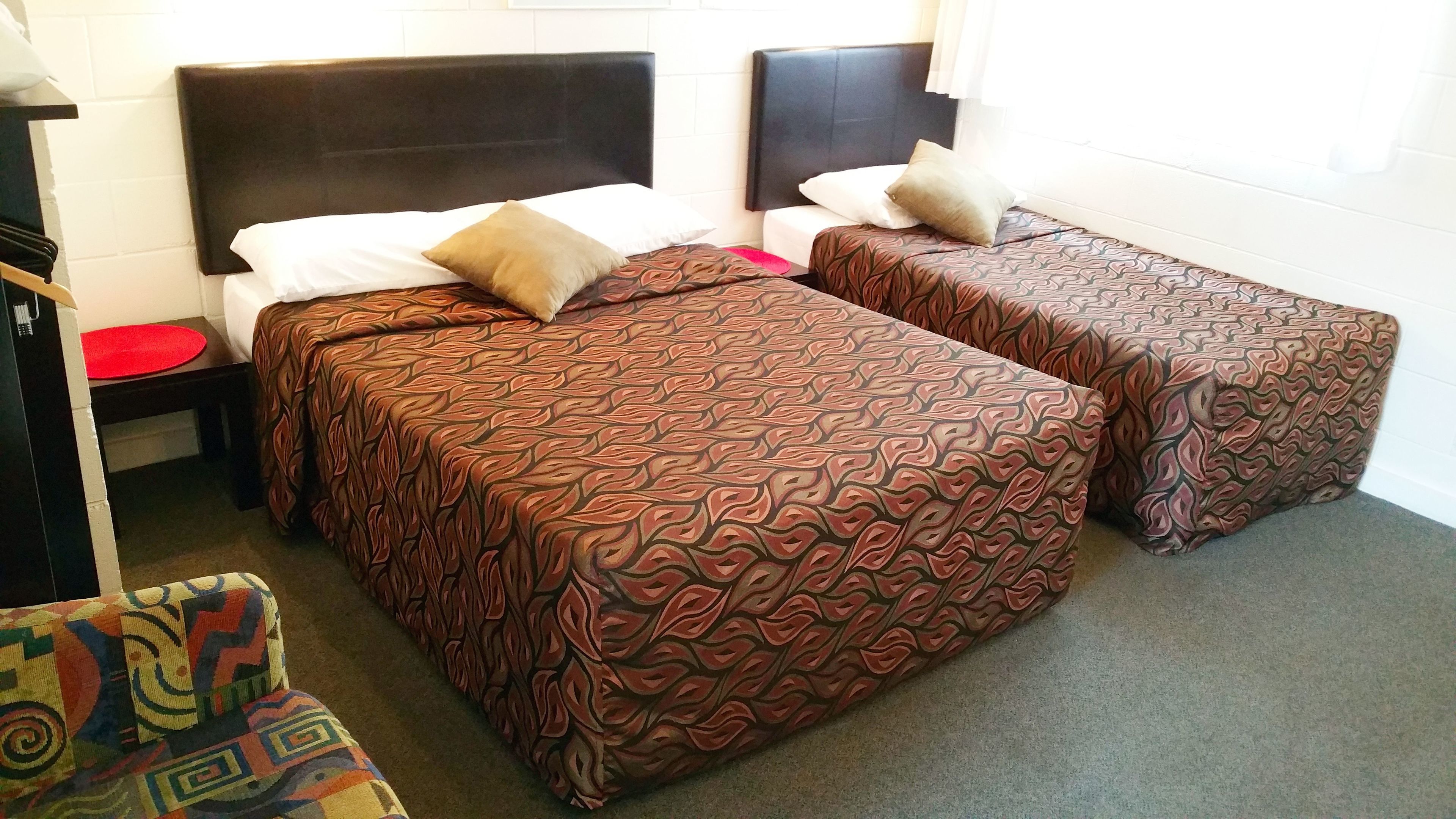 Quarto com duas camas, cada uma com colchas e almofadas estampadas, uma pequena mesa com um tapete vermelho e uma cadeira colorida.