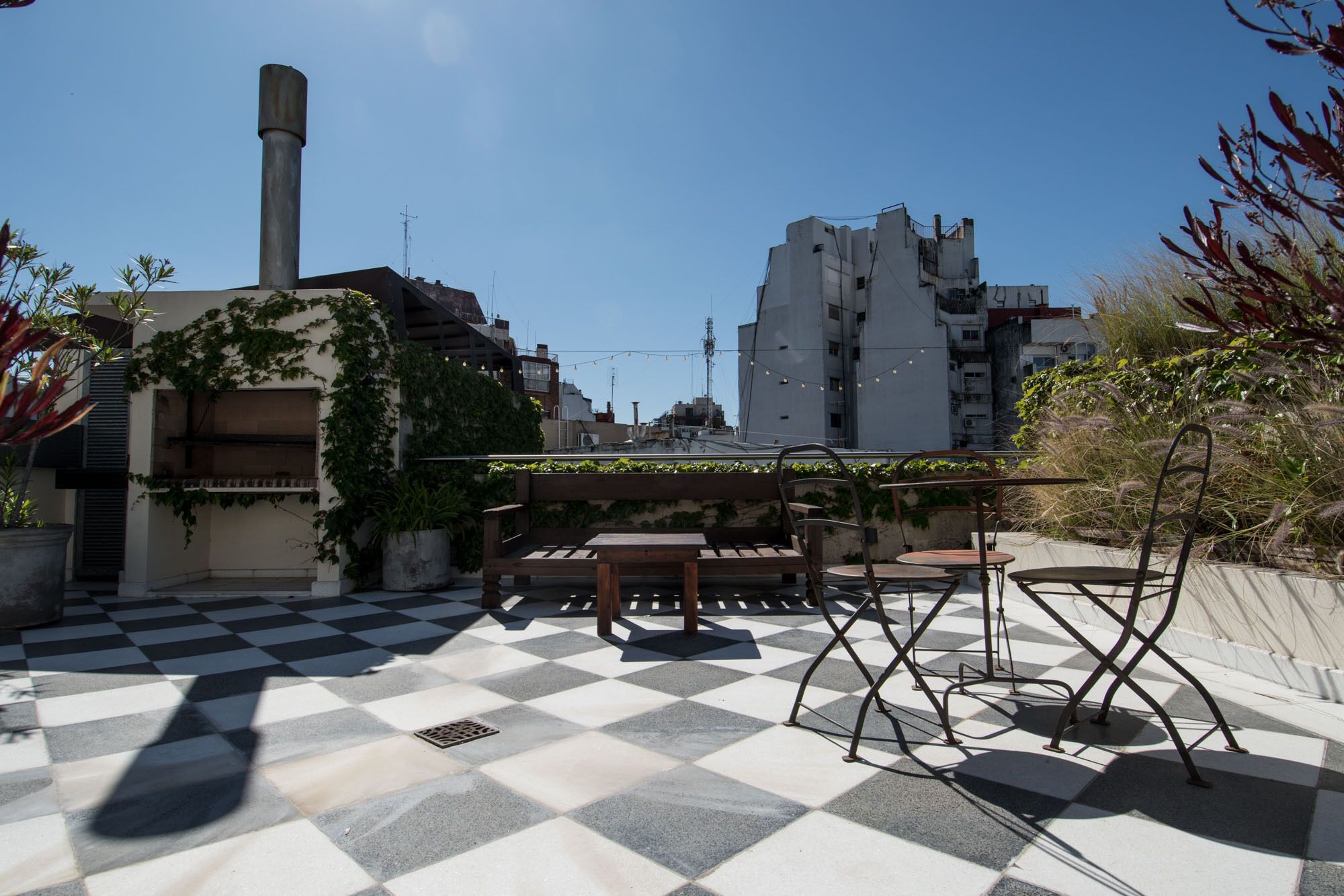 Terraço no telhado com piso xadrez, um banco de madeira e cadeiras de metal.