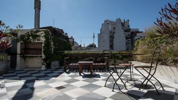 Terraço no telhado com piso xadrez, um banco de madeira e cadeiras de metal.