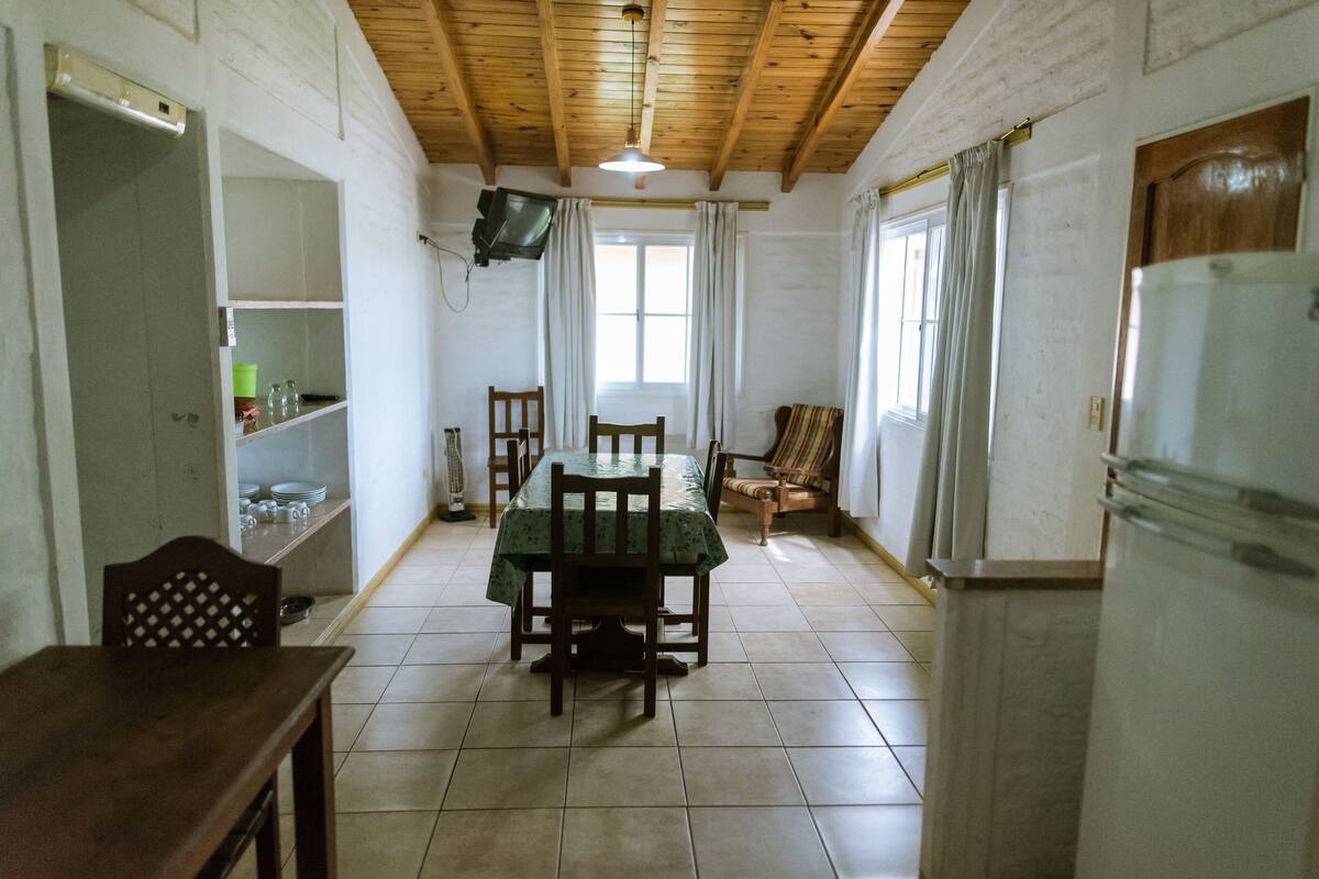 Uma sala de jantar com mesa e cadeiras de madeira, televisão na parede e um frigorífico.
