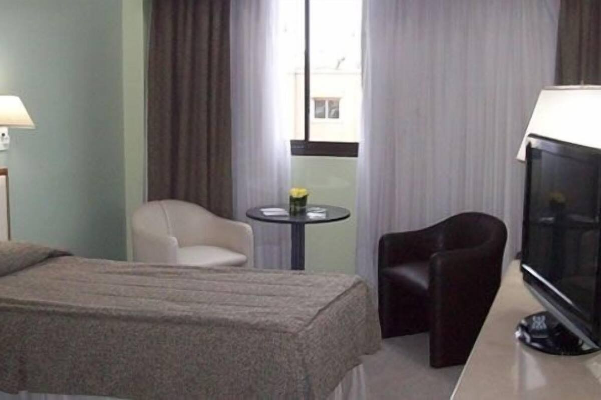 Quarto de hotel com cama, escrivaninha, cadeira, televisão e janela com cortinas.