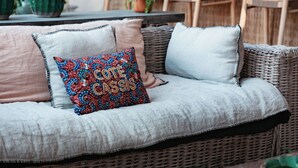 Um sofá de vime com almofadas estampadas, um espaço de bar com cadeiras e plantas em vasos.