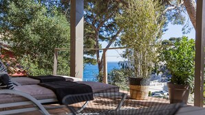 Uma área de estar ao ar livre e ensolarada com vista para o mar, incluindo uma cadeira de metal e uma mesa de vime com um vaso de planta.