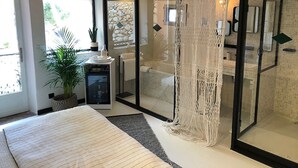 Quarto com cama, televisão, planta e porta de vidro deslizante que dá para um banheiro com chuveiro, pia e espelho.