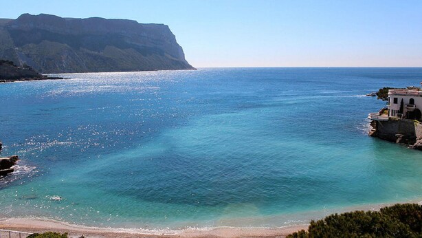 Cena costeira com mar azul límpido, linha de costa rochosa e um edifício branco com varanda.
