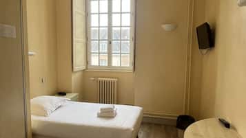 Quarto pequeno e compacto com uma cama de solteiro, uma mesa redonda, um radiador e uma janela com vista para prédios.