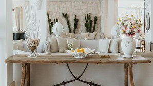 Uma mesa de madeira com um vaso de vidro, uma tigela de frutas e um vaso branco com flores em uma sala de estar moderna com parede de pedra e cactos.