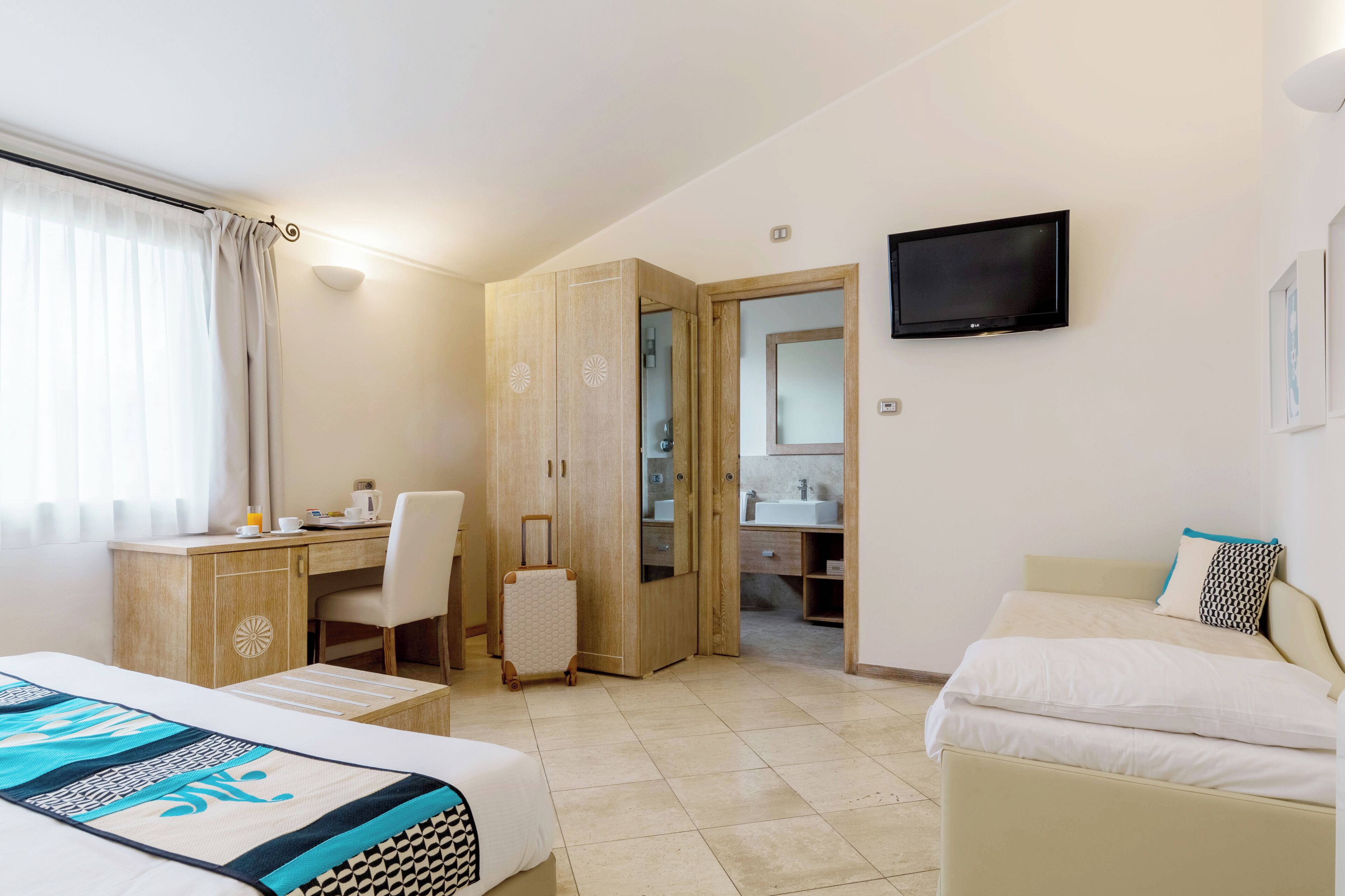 Quarto de hotel com cama, escrivaninha, cadeira, televisão e banheiro visível por uma porta aberta.
