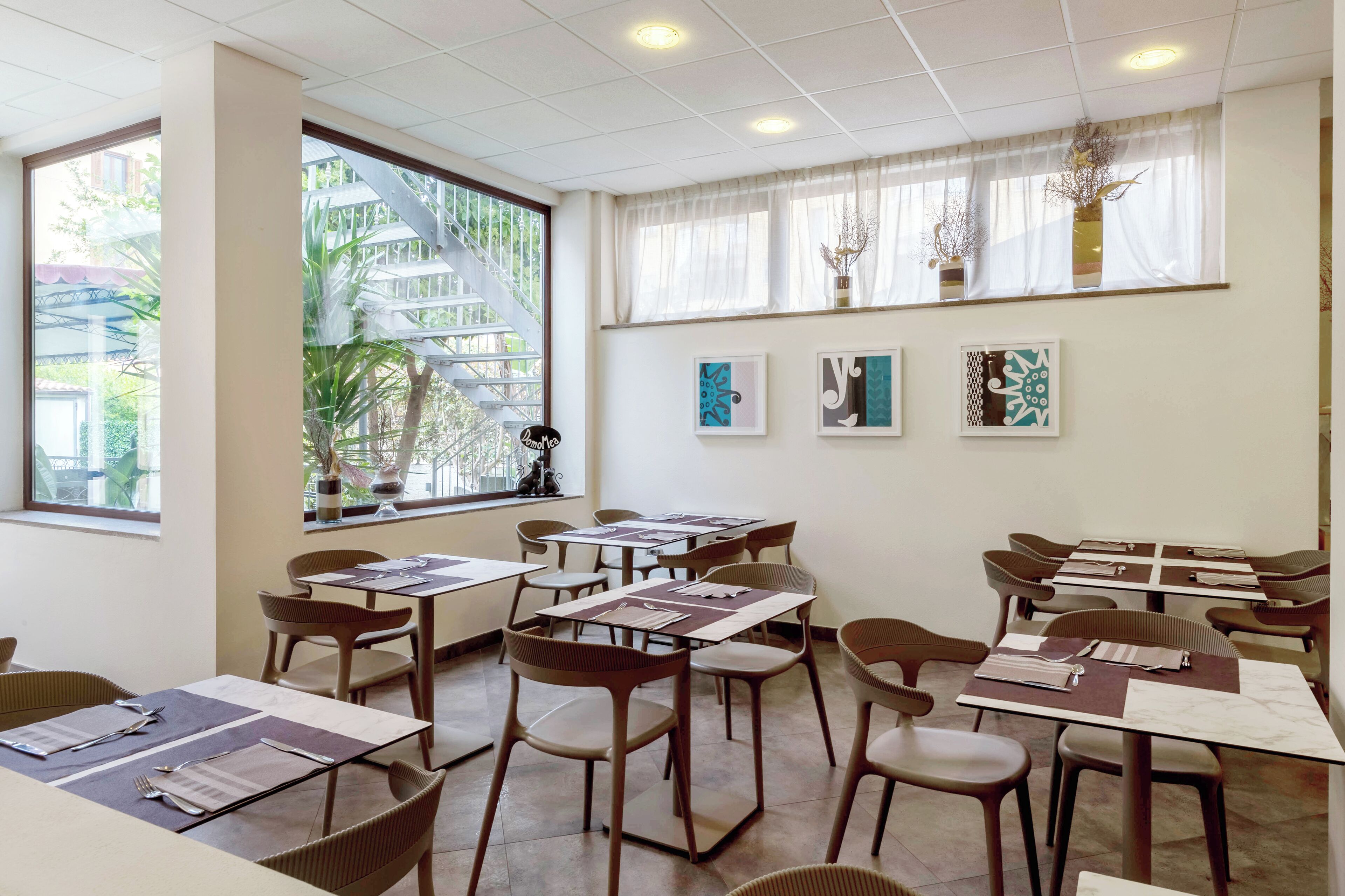 Uma sala de jantar com várias mesas e cadeiras, janelas amplas com cortinas e obras de arte nas paredes.