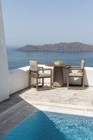 Uma varanda com piscina, duas cadeiras, uma mesa com uma tigela de fruta e vista para o mar e uma ilha ao longe.