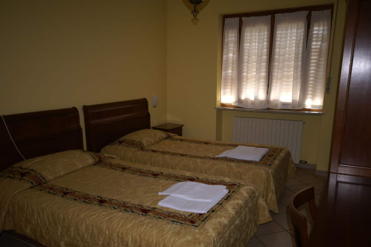 Quarto de hotel com duas camas, janela com cortinas, radiador e cabeceira de madeira.