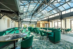 Restaurante com teto de vidro, assentos estofados em verde e estrutura central de madeira.