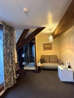 Quarto de hotel compacto com sofá cinza, uma pequena mesa com amenidades, cortina estampada, área de chuveiro em vidro e um vigamento de madeira no teto.