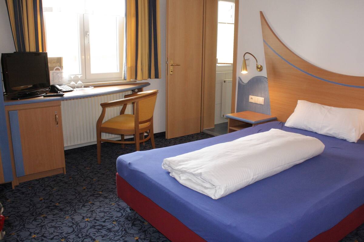 Quarto de hotel com cama, escrivaninha com computador, cadeira e mesa de cabeceira com abajur.