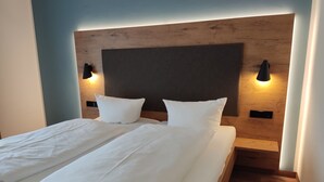 Cama de casal com lençóis brancos, cabeceira de madeira e duas luminárias de parede para leitura.