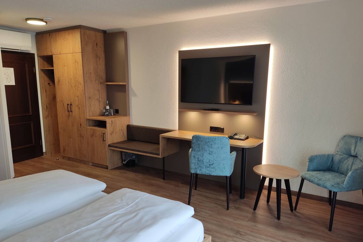 Quarto de hotel com cama, escrivaninha, cadeira, televisão e guarda-roupa.
