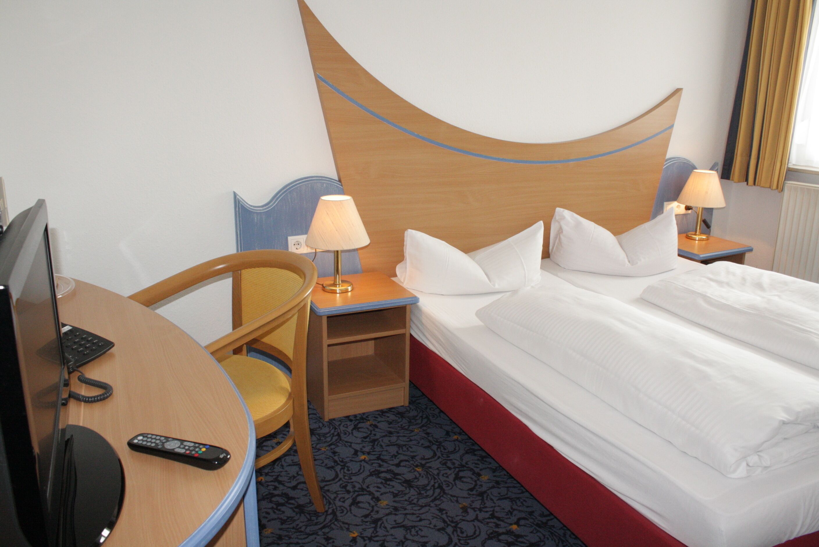 Quarto de hotel com cama, escrivaninha, computador, controle remoto, uma cadeira e duas luminárias de cabeceira.