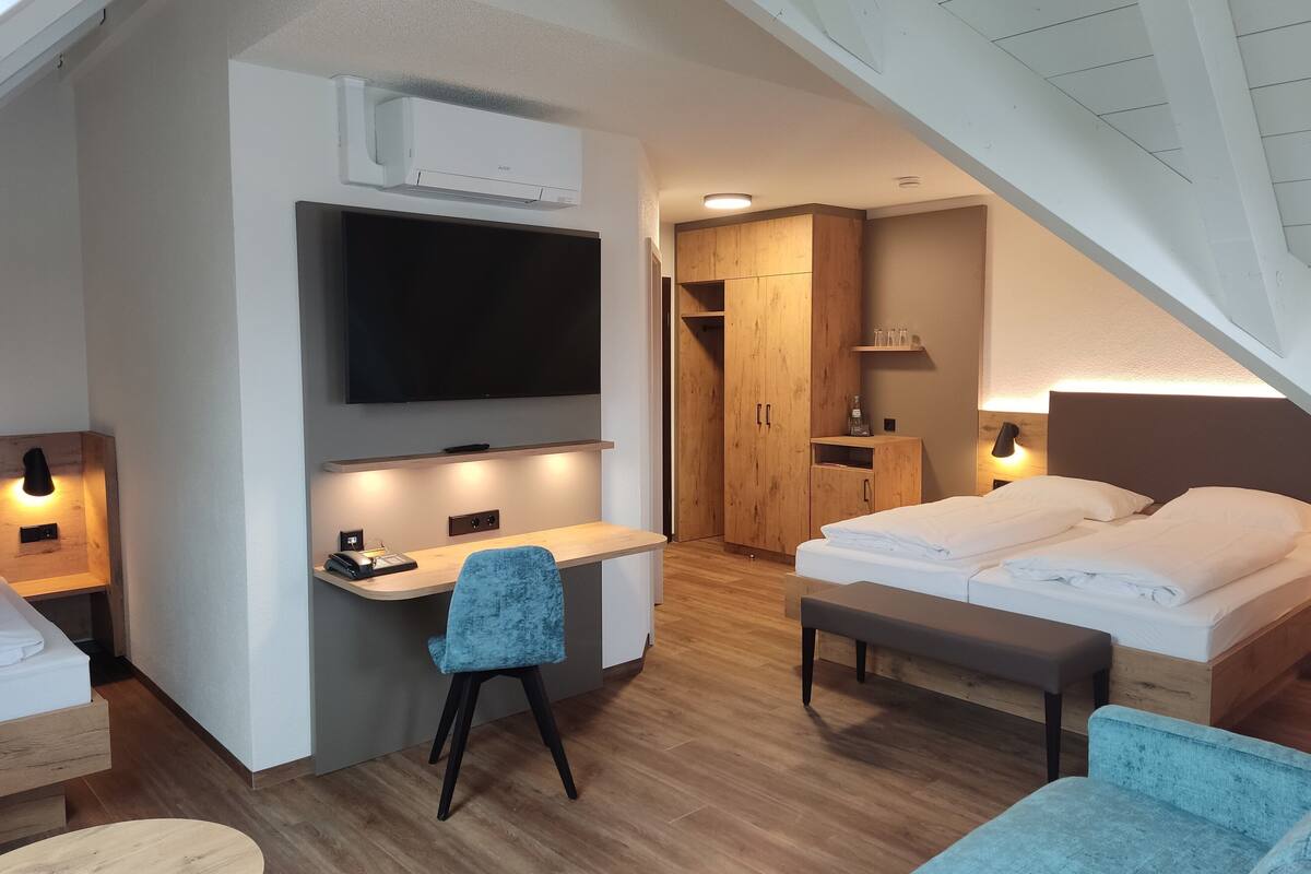 Quarto de hotel moderno com televisão de tela plana, escrivaninha com cadeira, cama com banco e guarda-roupa de madeira.
