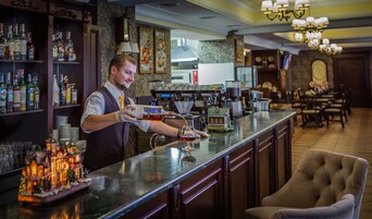 Uma área de bar com um bartender servindo uma bebida, prateleiras com garrafas de bebidas alcoólicas e um espaço de estar acolhedor com uma cadeira.