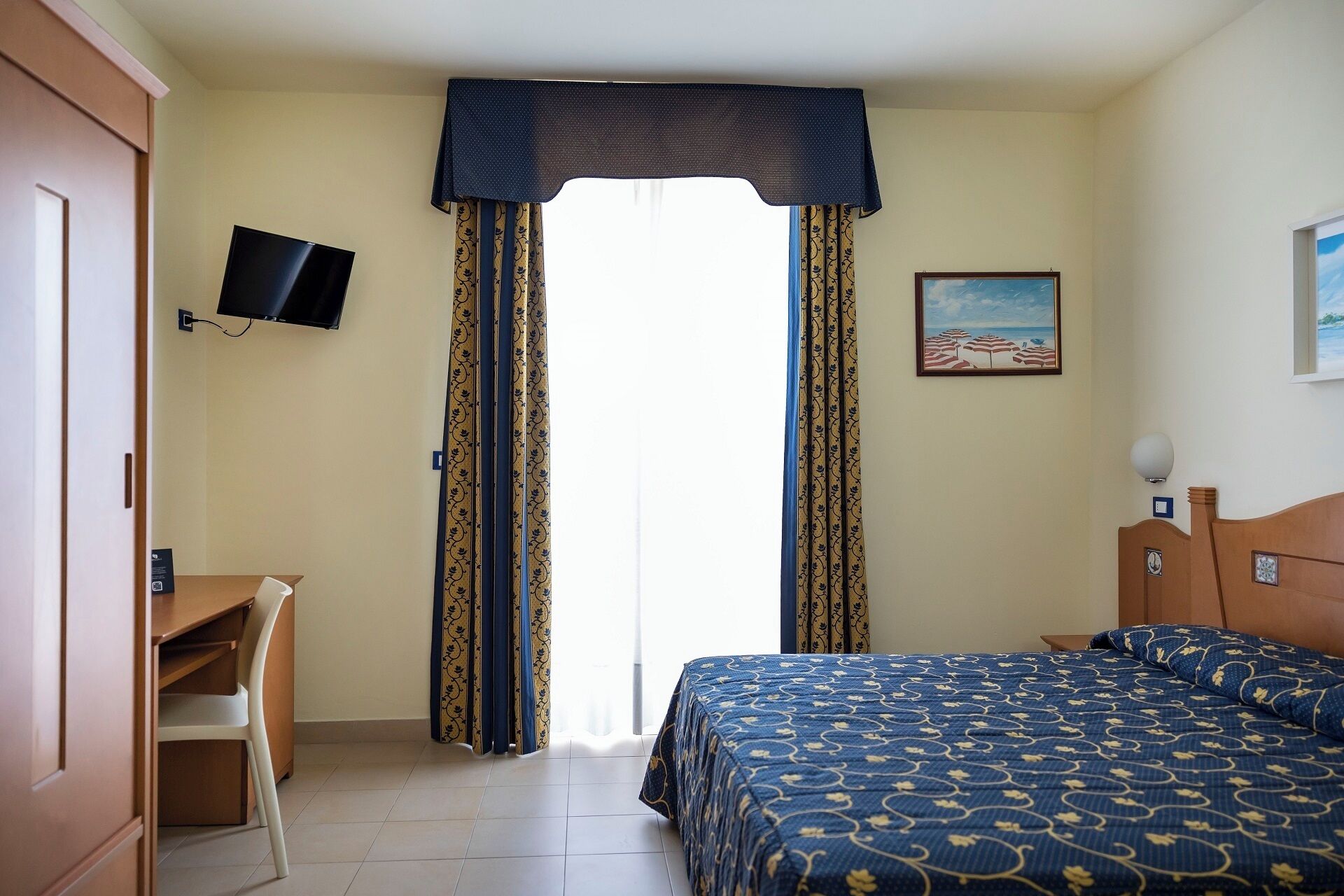 Quarto de hotel com cama, escrivaninha, televisão e cortinas.