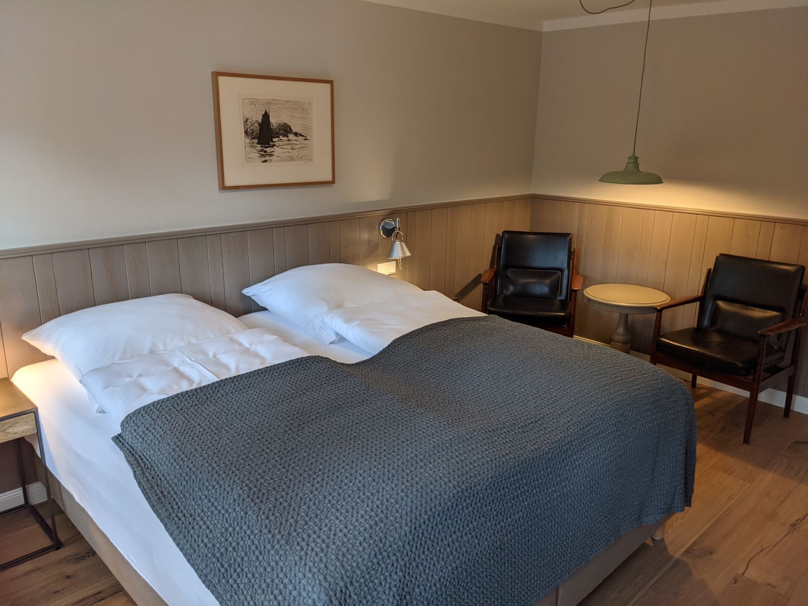 Uma cama bem arrumada com lençóis brancos e um cobertor cinza, uma obra de arte emoldurada na parede, uma luminária pendente verde e duas poltronas de couro.