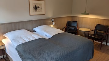 Uma cama bem arrumada com lençóis brancos e um cobertor cinza, uma obra de arte emoldurada na parede, uma luminária pendente verde e duas poltronas de couro.