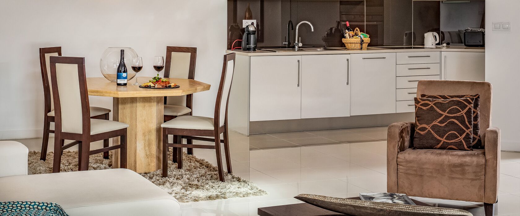 Uma cozinha moderna com armários brancos, uma área de refeições com mesa e cadeiras de madeira, e um espaço de estar aconchegante com uma poltrona marrom e um sofá branco.