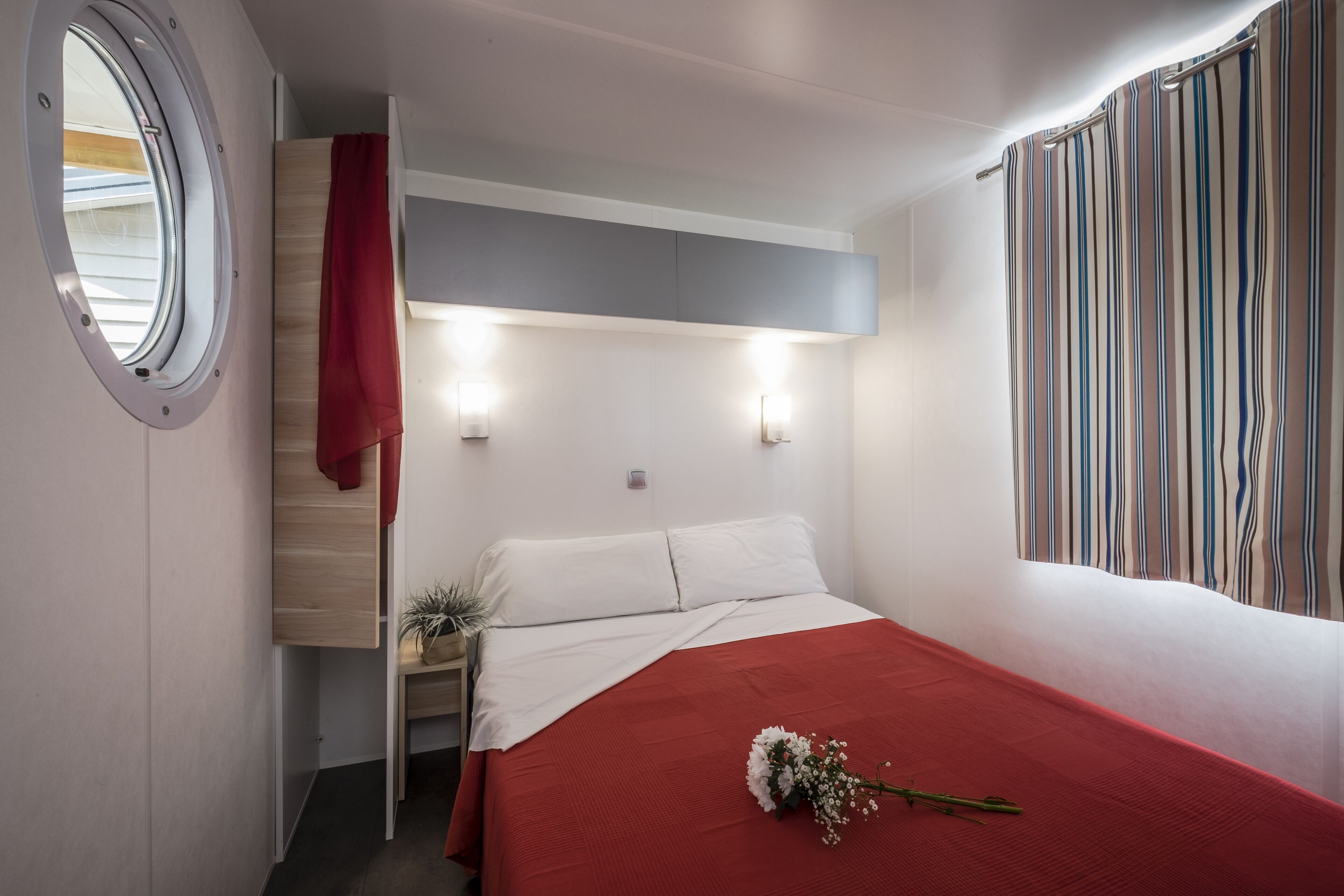 Quarto com cama, janela, cortina vermelha, uma pequena planta e luminária fixada na parede.