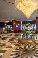 Um hall de entrada com um lustre grande, uma mesa redonda dourada com arranjos florais e um chão quadriculado.
