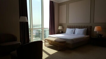 Quarto de hotel com uma cama grande, uma mesa pequena, uma cadeira e vista da cidade através de janelas amplas.