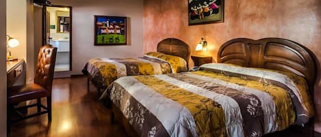 Quarto de hotel com chão de madeira, duas camas com roupa de cama floral, cabeceira de madeira, uma escrivaninha com cadeira e um quadro na parede.