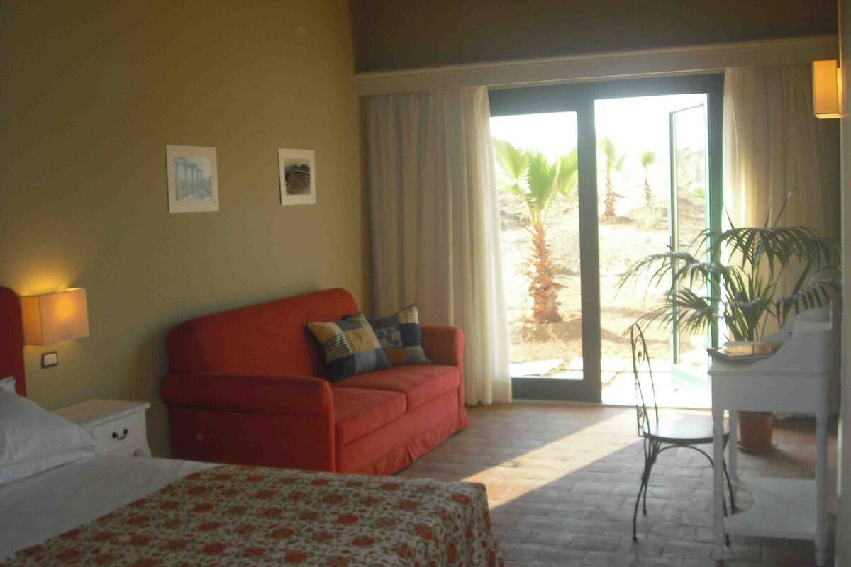 Quarto de hotel com uma cama, um sofá vermelho, uma mesinha com uma planta e vista para palmeiras pela porta.