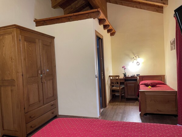 Quarto com armário de madeira, cama com colcha vermelha, escrivaninha com cadeira e mesa de cabeceira com abajur.