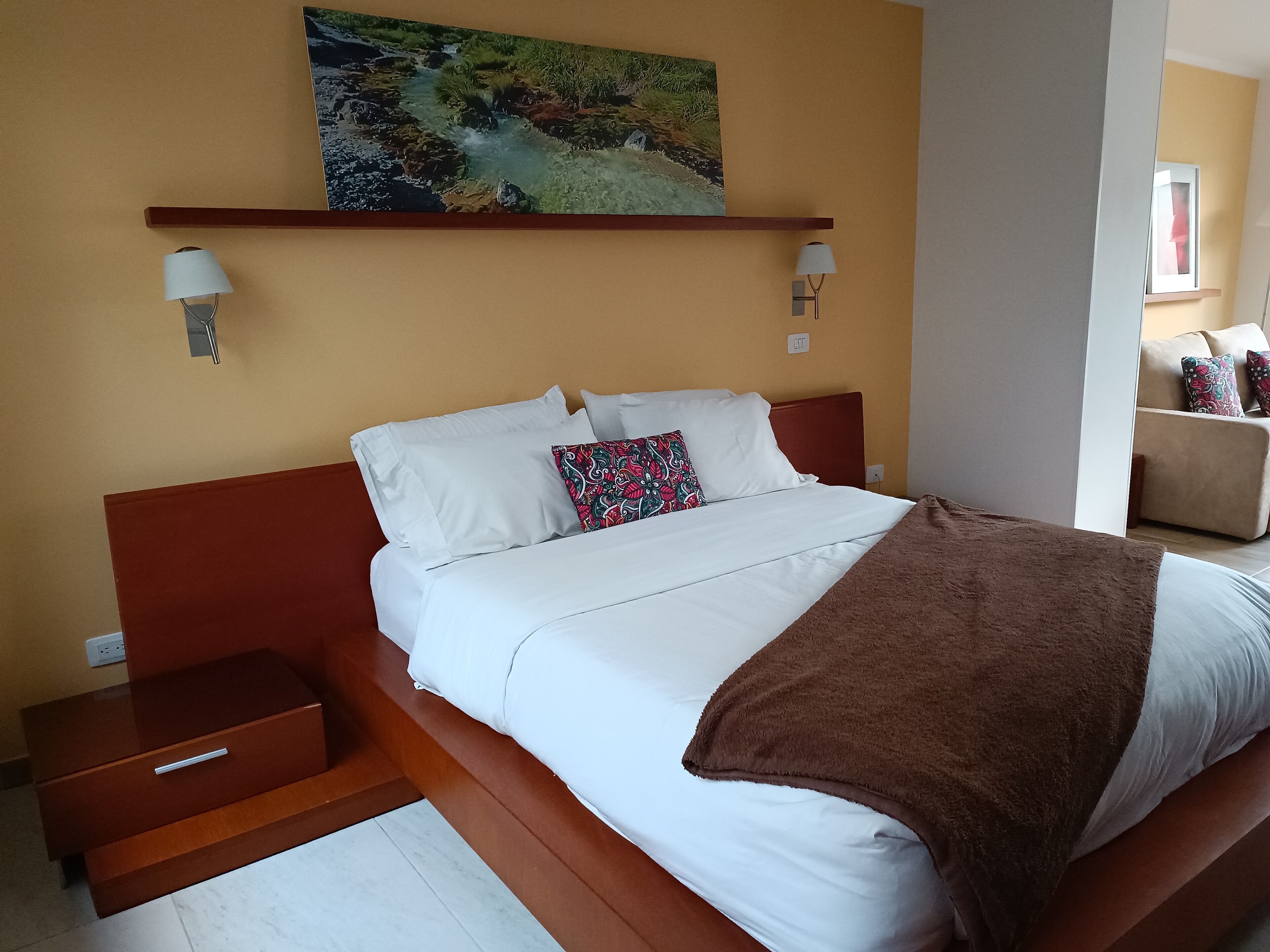Cama bem arrumada com lençóis brancos e um cobertor marrom, mesa de cabeceira de madeira com gaveta, pintura de uma paisagem fluvial na parede e um sofá com almofadas estampadas ao fundo.