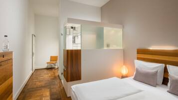 Quarto de hotel com piso de madeira, cama com roupa de cama branca, mesa de cabeceira com abajur e espelho na parede.