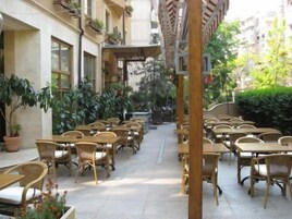 Uma área de café ao ar livre com mesas e cadeiras de vime, plantas em vasos e uma cobertura.