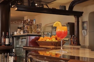 Uma área de bar com um copo de drink vermelho decorado com uma fatia de laranja, uma cesta de laranjas e diversas garrafas de álcool nas prateleiras.
