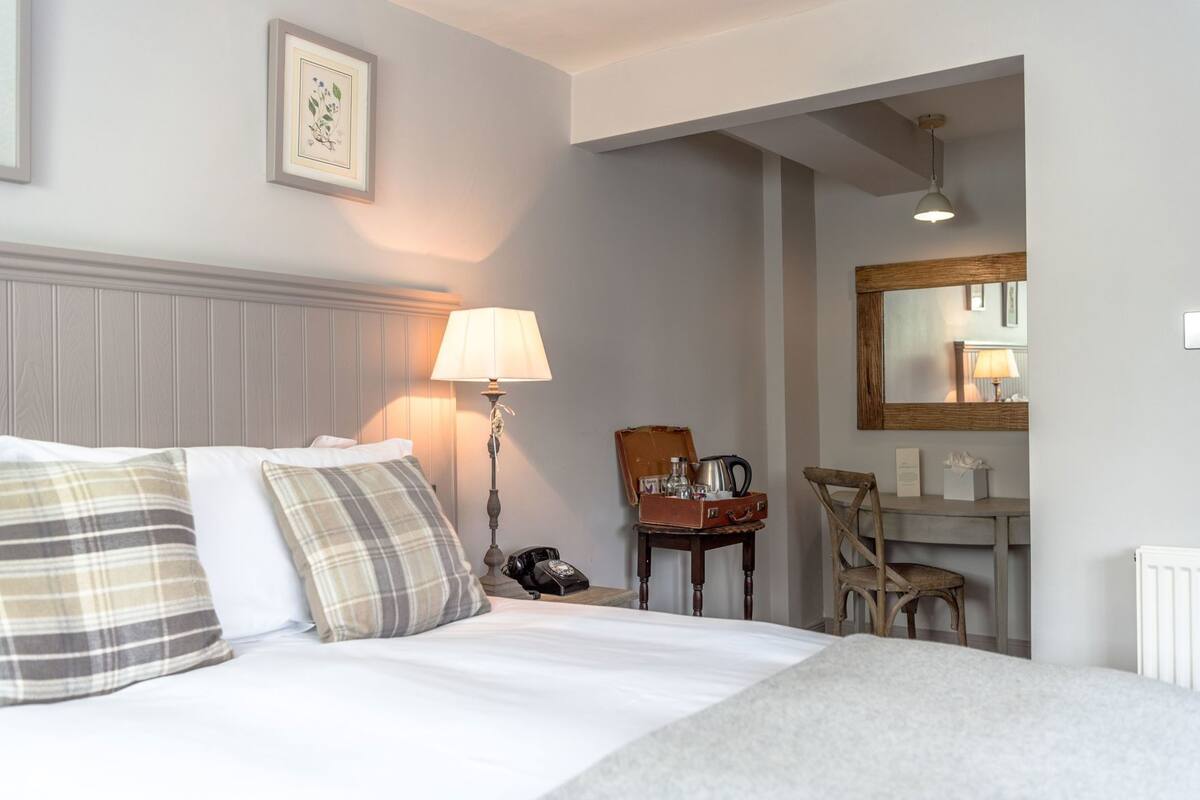 Quarto de hotel com cama, escrivaninha, cadeira, luminária, telefone e espelho.