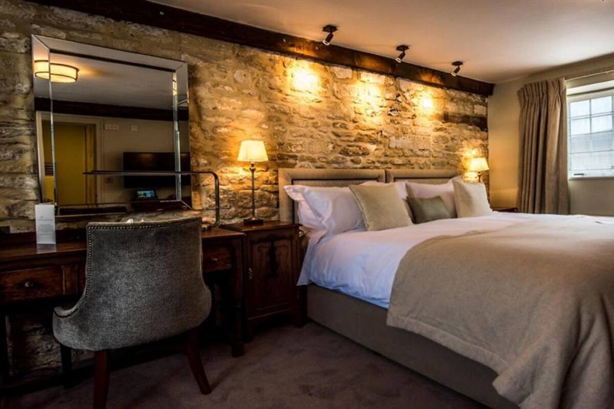 Quarto com parede de pedra, cama com roupa de cama branca, escrivaninha com cadeira e duas luminárias de cabeceira.