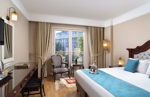 Quarto de hotel com cama, escrivaninha, cadeira, janela com cortinas e mesa de cabeceira com abajur.