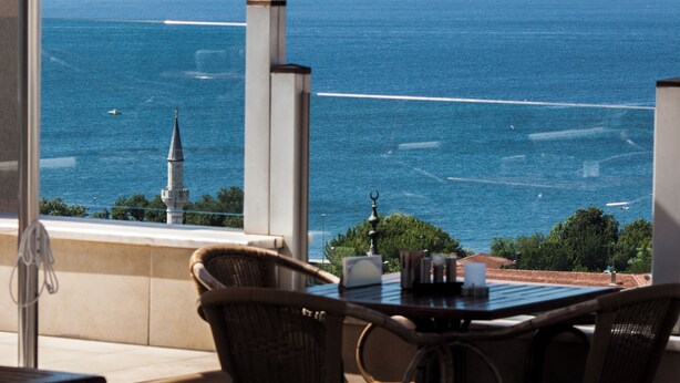 Um terraço à beira-mar com cadeiras de vime e uma mesa, com vista para o oceano e um navio ao longe.