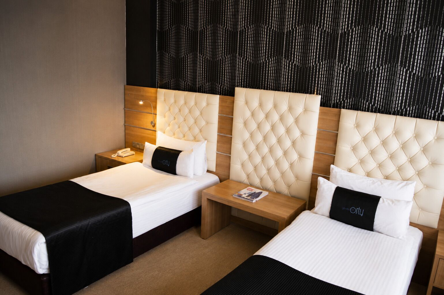 Quarto de hotel com duas camas, uma cabeceira de madeira, uma mesa de cabeceira com telefone e uma parede com textura.